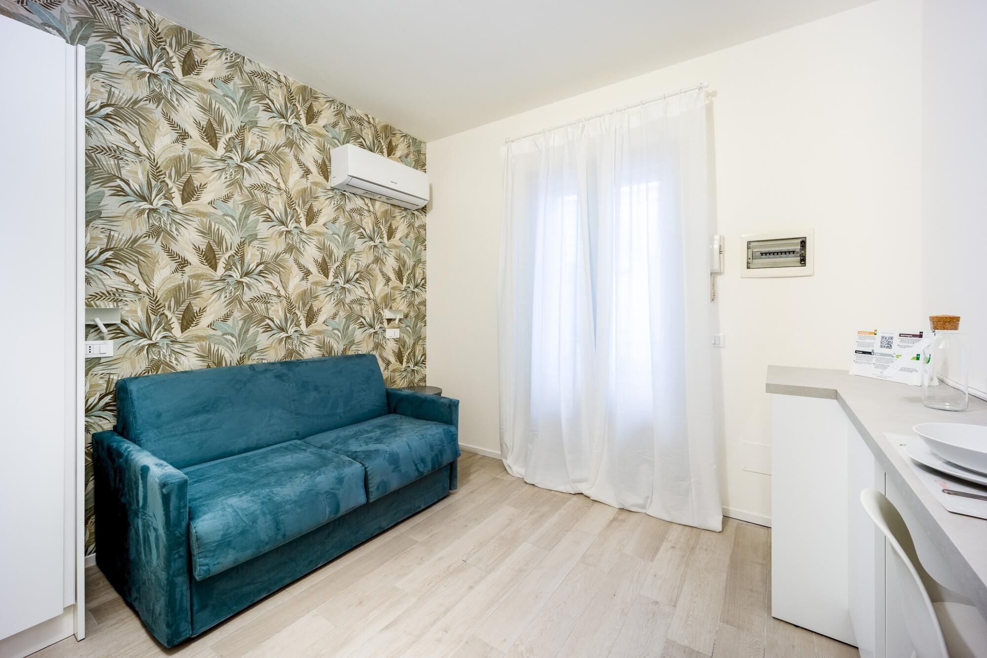 Quarto com um sofá azul, um ar condicionado branco, uma cortina branca, um pequeno armário branco e uma parede com padrão de folha tropical.