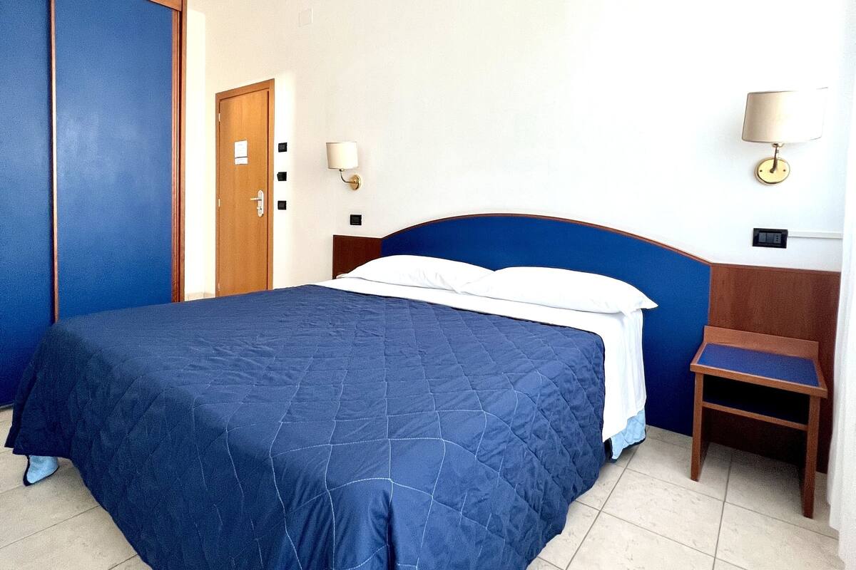 Quarto de hotel com cama, mesa de cabeceira, luminária, ventilador de teto e ar condicionado.