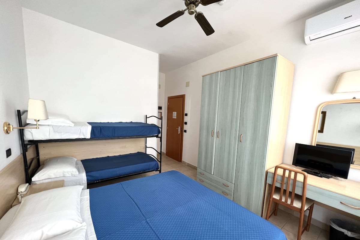 Quarto de hotel com beliche, escrivaninha com cadeira, laptop, ventoinha no teto e guarda-roupa.