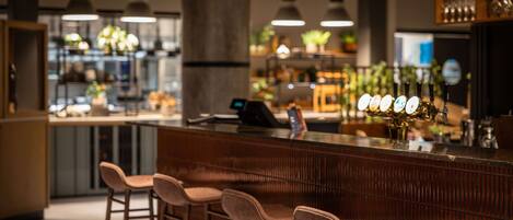 Uma área de bar com banquetas, balcão com caixa registradora e copos pendurados.