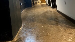 Um corredor com piso de concreto, parede preta à esquerda e parede branca à direita. Há duas portas à direita, sendo uma delas aberta.