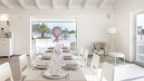 Uma área de refeições com uma mesa comprida posta para uma refeição, cadeiras brancas, uma pequena mesa lateral com uma luminária e vista para o exterior através de uma grande janela.