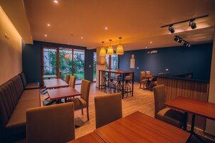 Restaurante com mesas e cadeiras de madeira, área de bar e grandes janelas com vista para o verde lá fora.