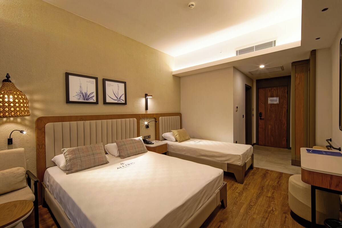 Quarto de hotel com duas camas, uma escrivaninha, uma cadeira, um abajur e impressões botânicas emolduradas na parede.