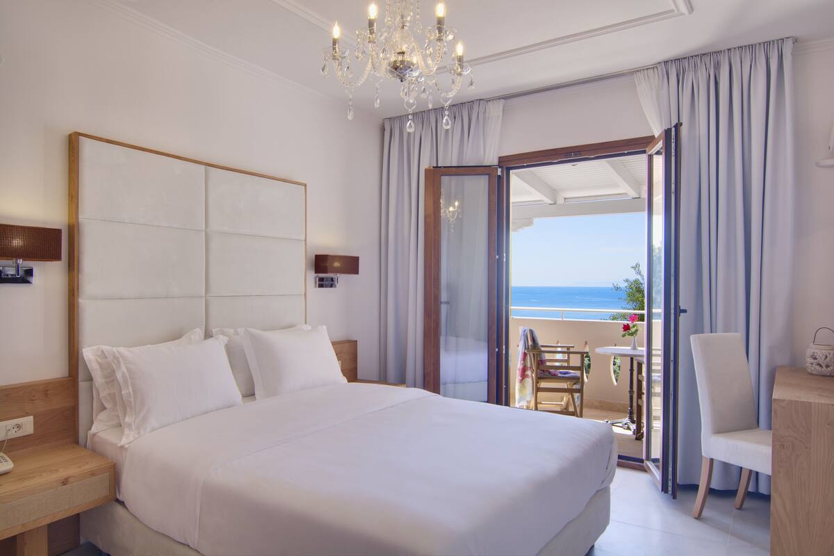 Quarto de hotel com uma cama grande, um candelabro, uma varanda com vista para o mar e uma pequena mesa com cadeiras.
