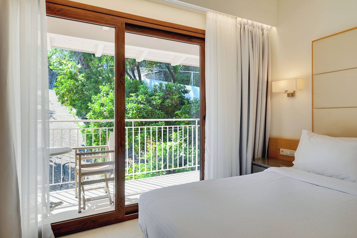 Quarto de hotel com cama, mesa de cabeceira, abajur, janela com cortinas e vista para uma varanda com vegetação.