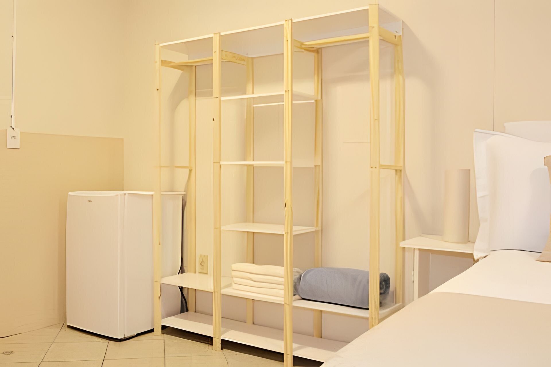 Quarto com cama, guarda-roupa com várias prateleiras, um frigorífico pequeno e branco e janela com cortina.