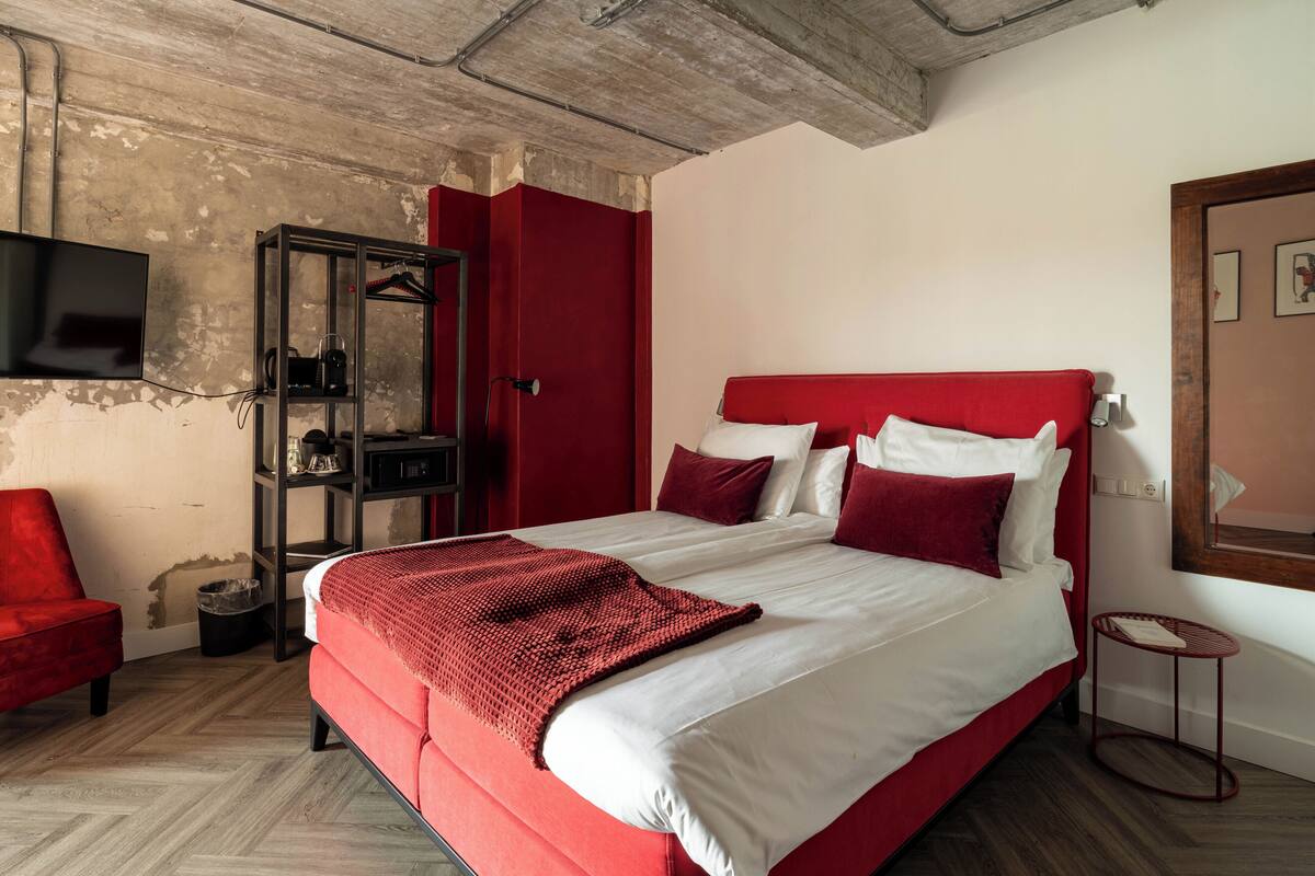 Quarto de hotel com uma cama grande, cabeceira vermelha, televisão na parede, uma cadeira vermelha e uma mesa pequena.