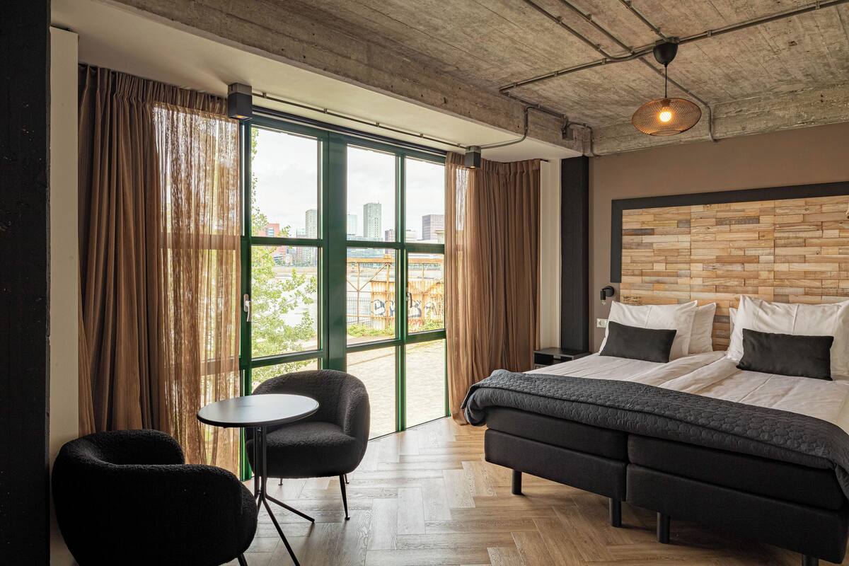 Quarto de hotel moderno com uma cama grande, uma mesa redonda pequena, duas cadeiras pretas e vista para a cidade através de amplas janelas.