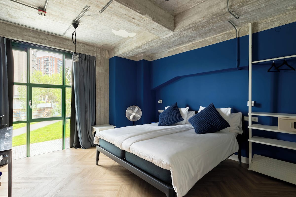 Quarto com uma cama grande, uma mesinha de cabeceira, um ventilador e vista para um edifício pela janela.