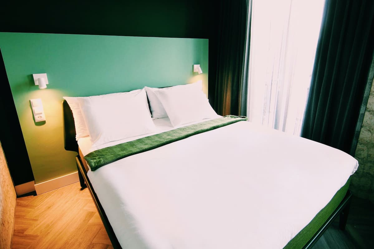 Cama bem arrumada com lençóis brancos e colcha verde em quarto com paredes verde-água, piso de madeira e janela com cortinas.