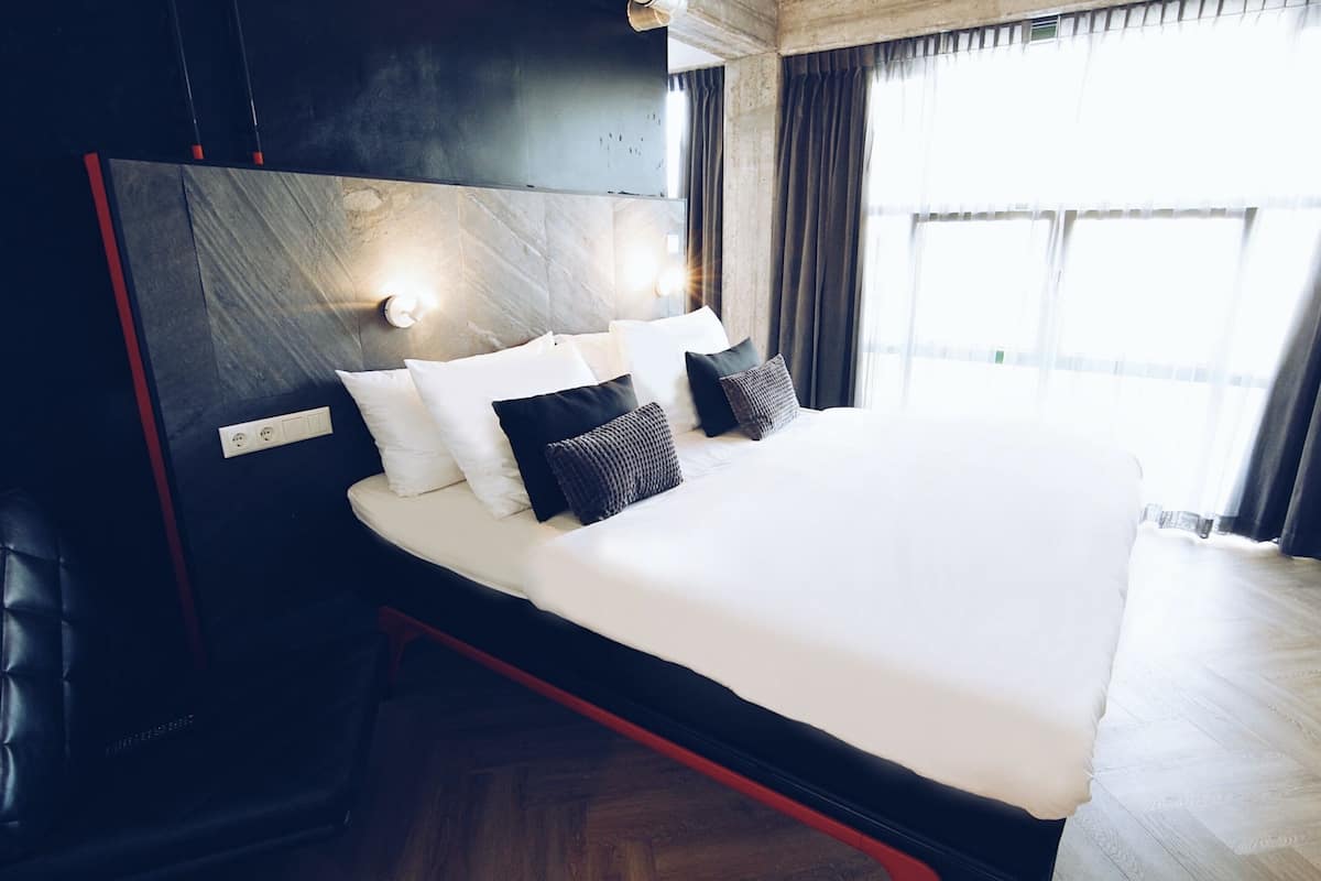 Quarto de hotel moderno com cama grande, cabeceira escura e vista para a cidade através de janelas amplas.