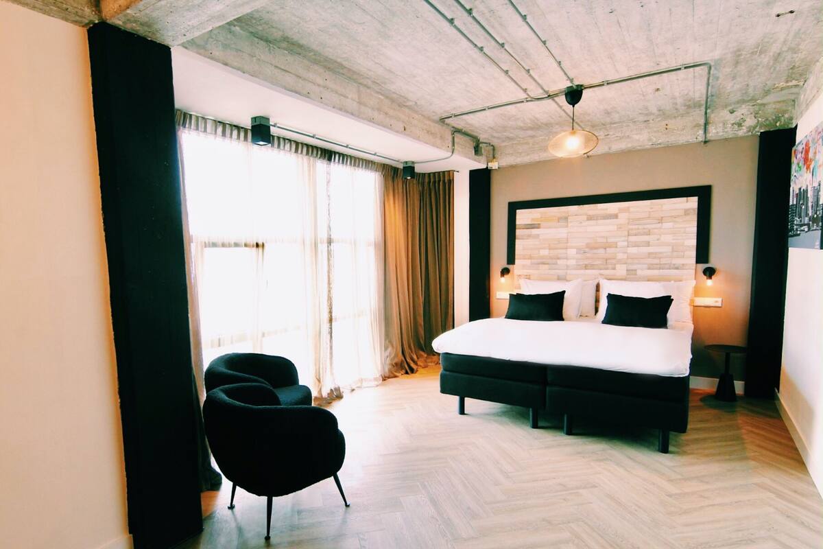Quarto de hotel moderno com uma cama grande, uma cadeira preta e um piso de madeira em padrão espinha de peixe.