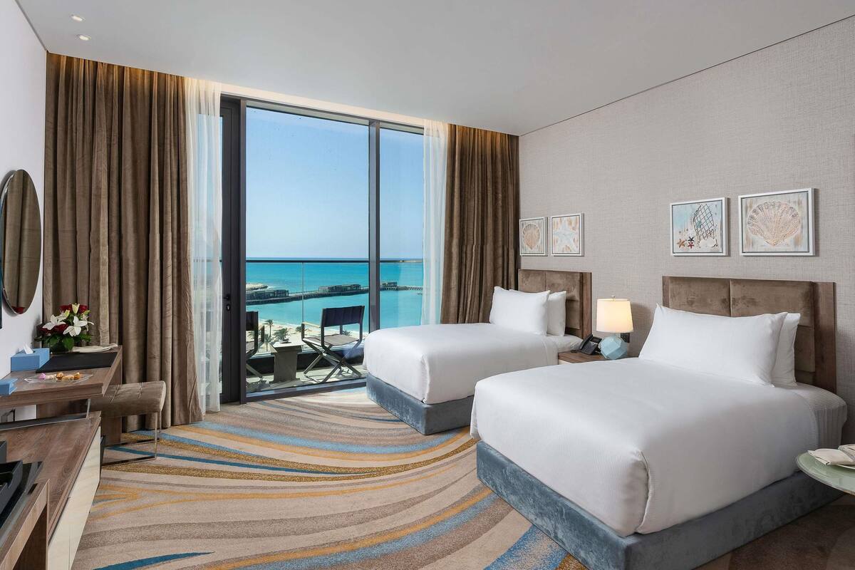 Quarto de hotel com duas camas, uma janela grande com vista para o mar, uma escrivaninha com cadeira e uma pequena varanda.