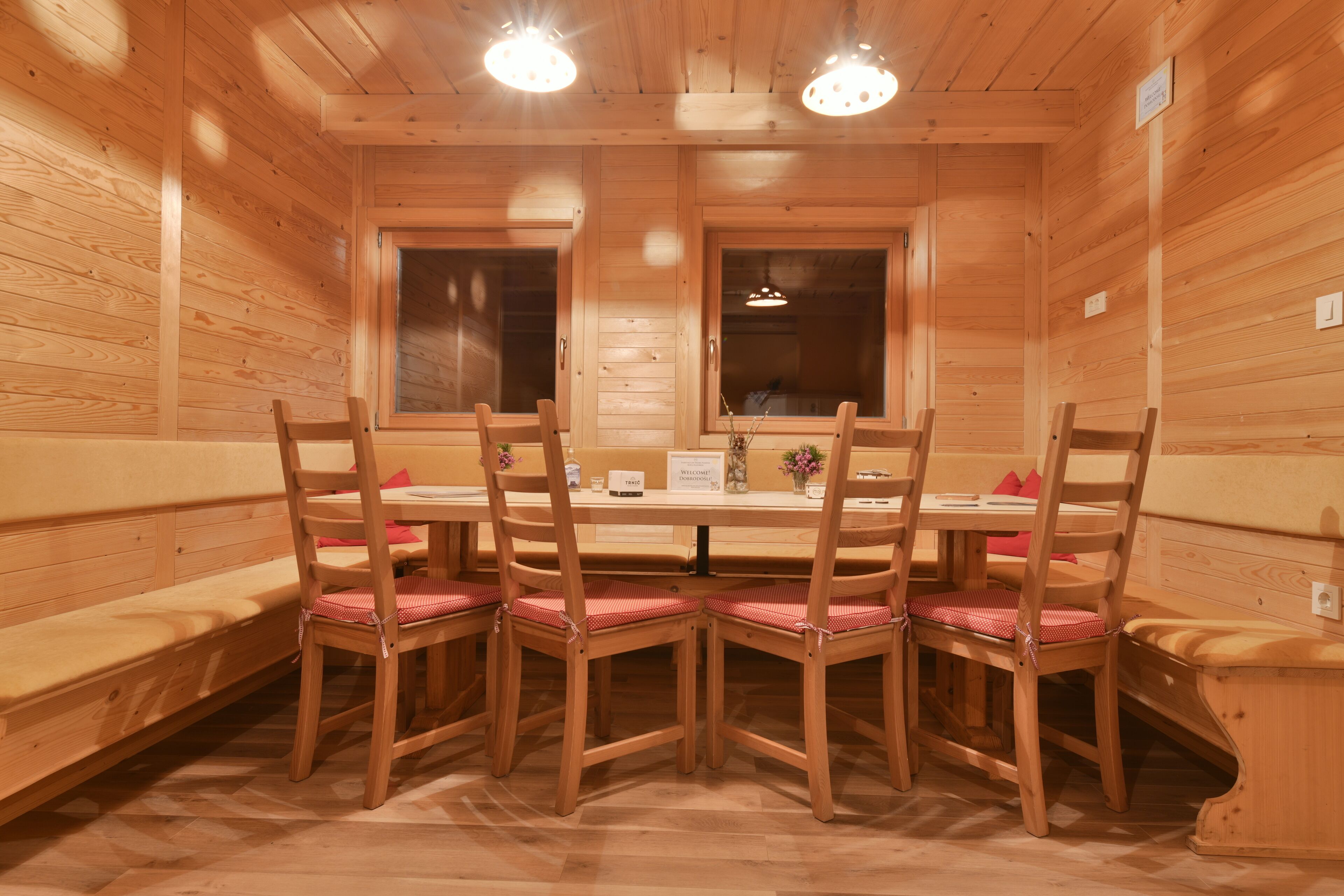 Interior de uma cabana de madeira com uma mesa de jantar cercada por cadeiras de madeira com almofadas vermelhas, um banco ao longo da parede e duas janelas.