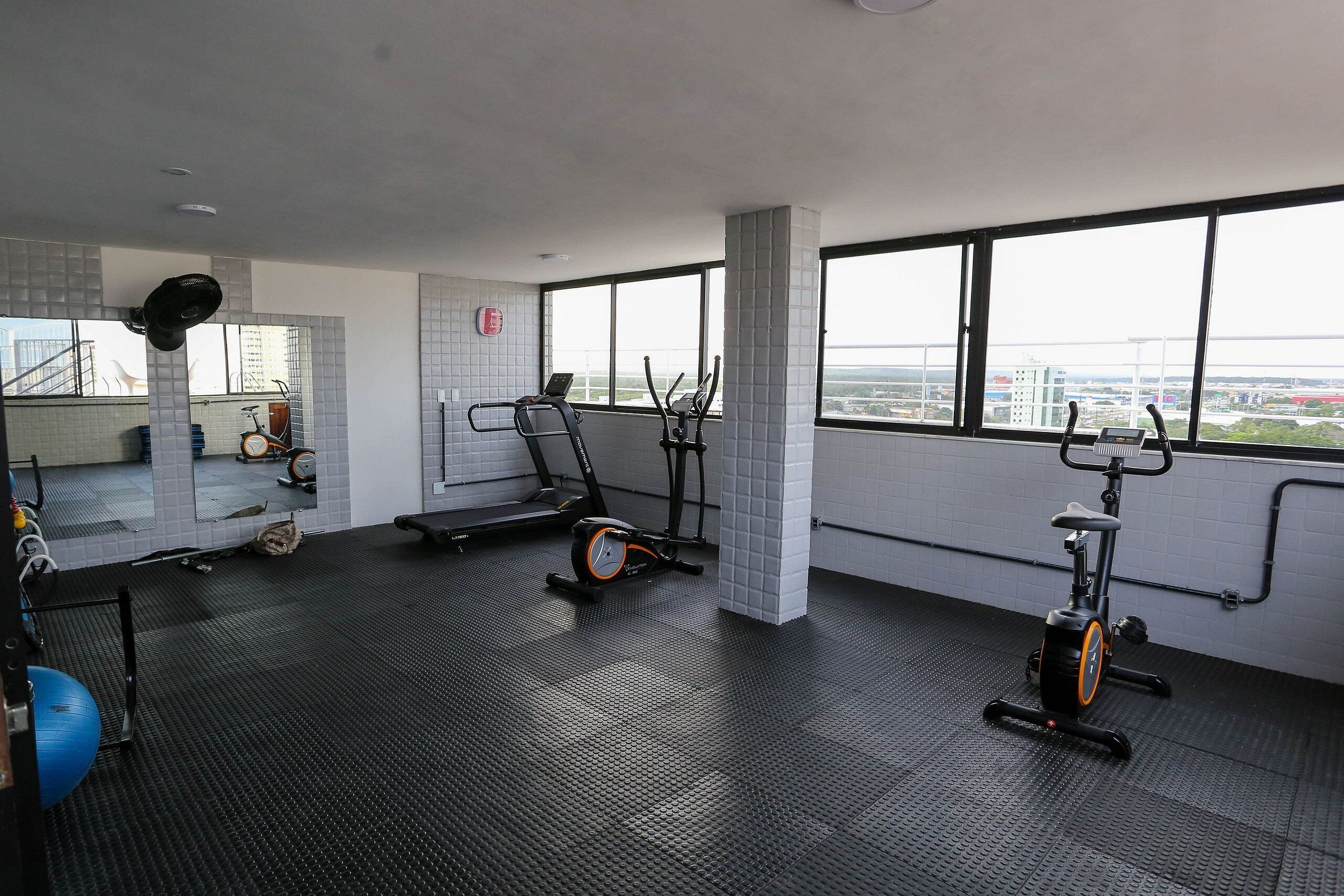 Academia com equipamentos de ginástica, janelas amplas e vista da cidade.