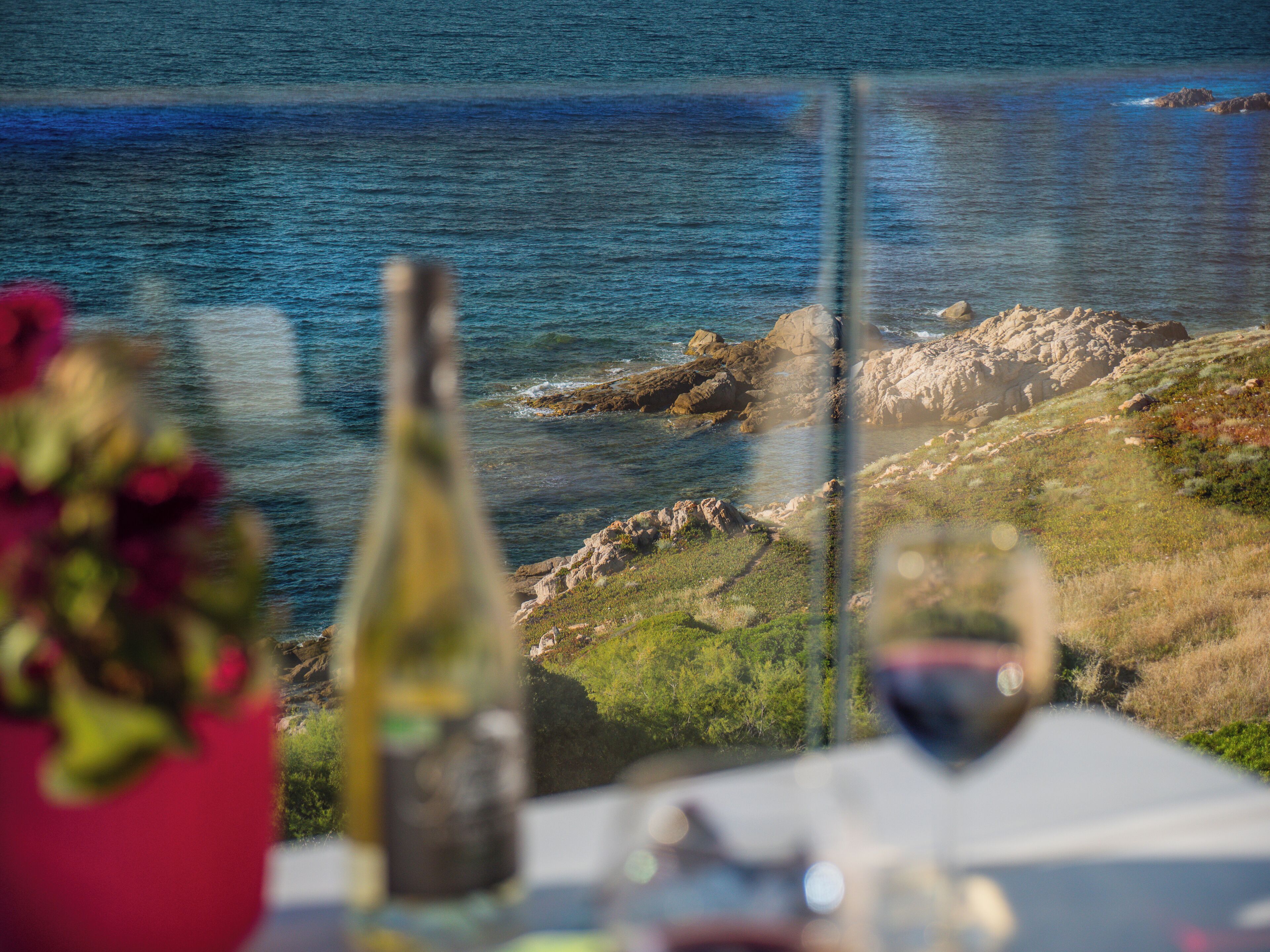 Uma mesa com uma garrafa e um copo, com vista para uma paisagem costeira com rochas e vegetação.