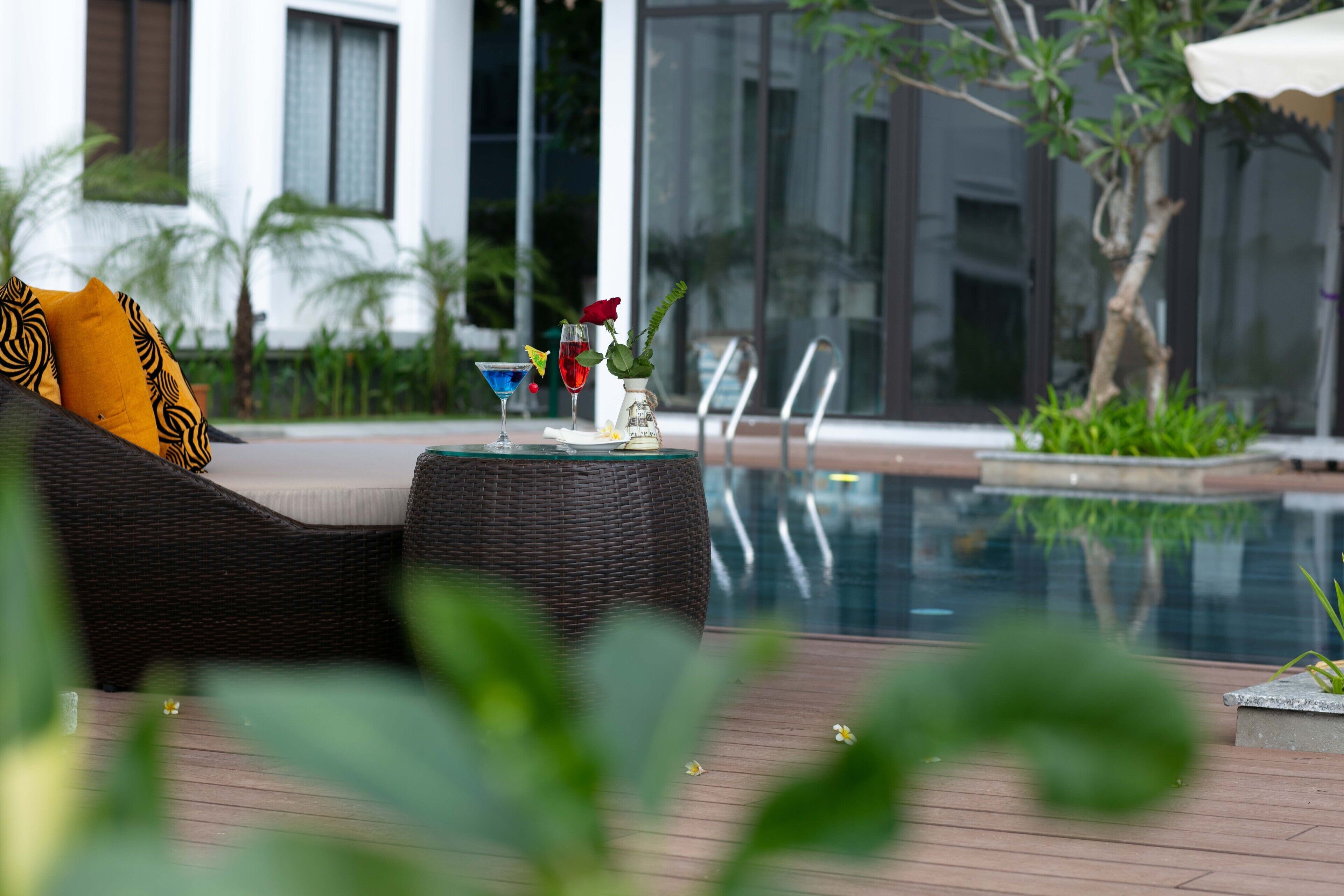Área ao redor da piscina com móveis de vime, uma mesa de vidro com bebidas e vista para a piscina e o edifício.