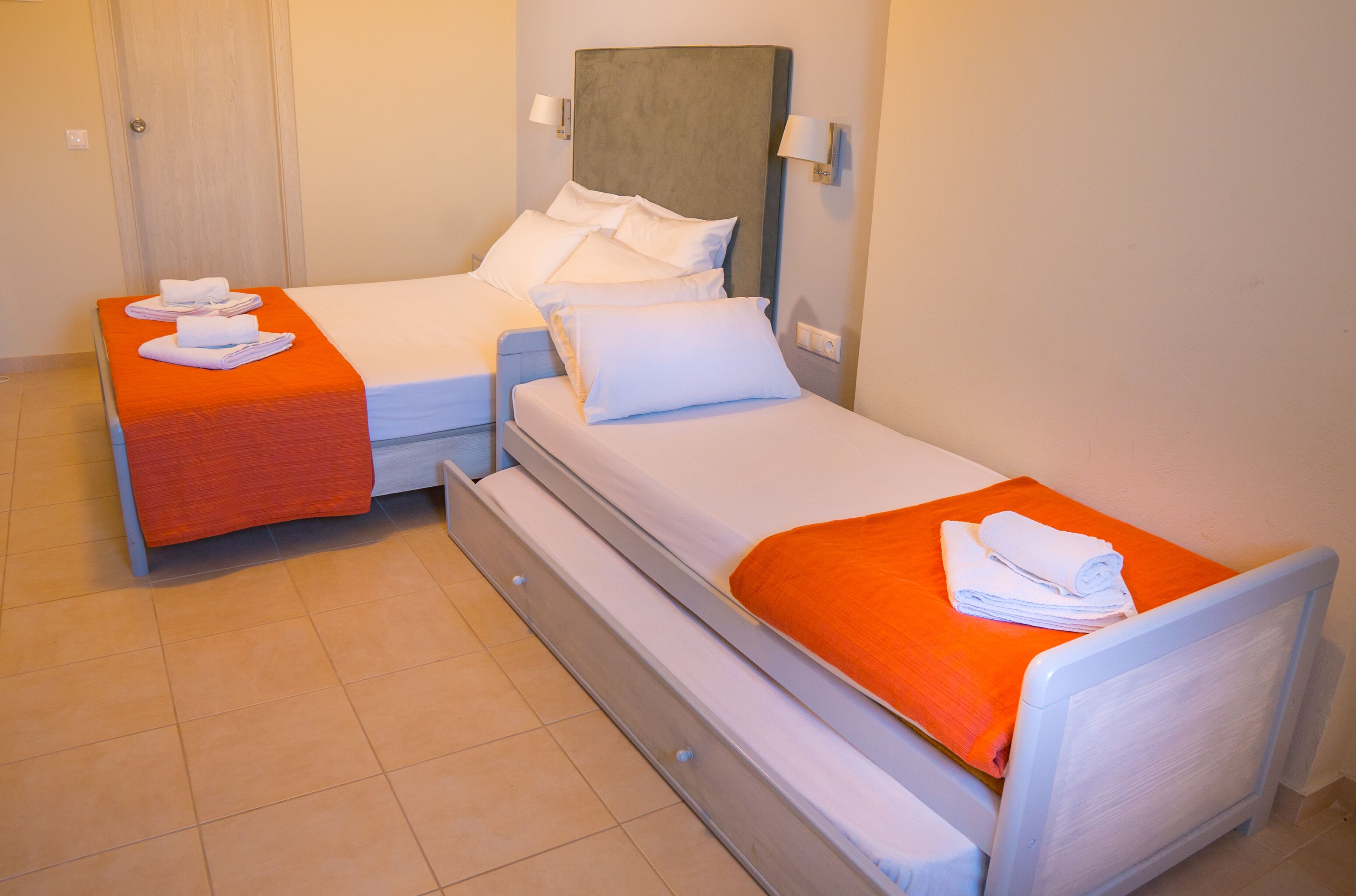 Quarto de hotel com duas camas, cada uma com colcha laranja e roupa de cama branca. Há duas luminárias fixadas na parede, uma de cada lado das camas.