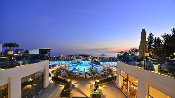 Uma área de piscina no terraço com espreguiçadeiras e guarda-sóis, cercada por edifícios e vista para o mar.