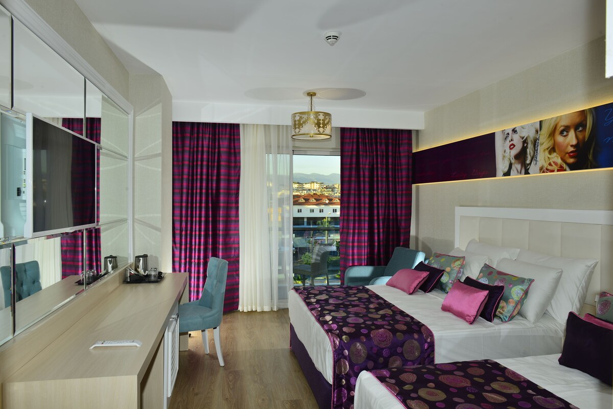 Quarto de hotel com cama, escrivaninha e cadeira. Janela com cortinas, ventilador de teto e vista da cidade.