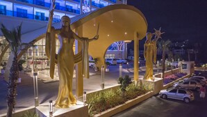Vista noturna da entrada de um hotel, com estátuas douradas de mulheres, uma galeria coberta e um estacionamento com carros.