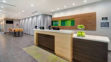 Uma área de recepção de hotel com design moderno, incluindo um balcão de recepção, um posto de check-in, um espaço de estar com cadeiras e uma placa verde 'HOME2' na parede.