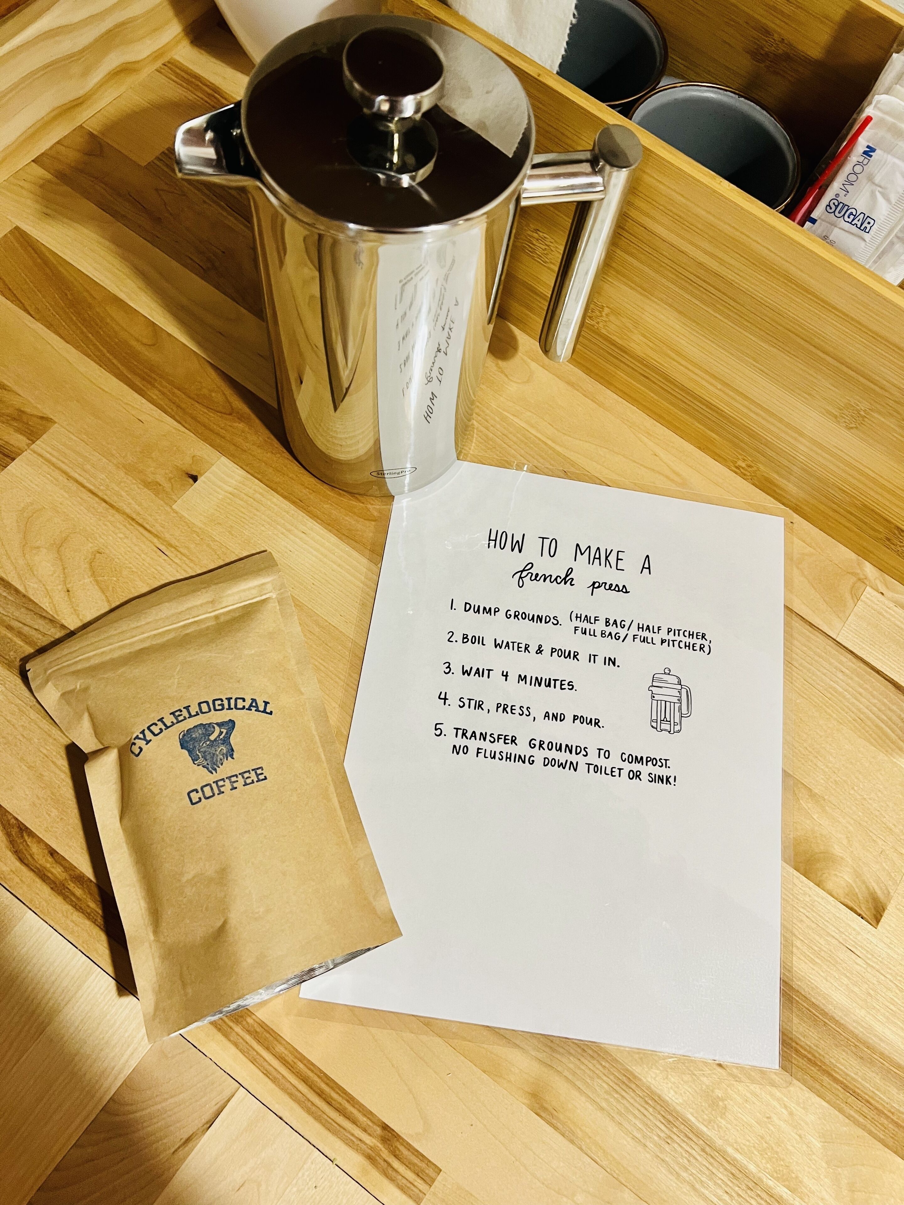 Uma cafeteira, um papel com instruções para fazer um furador e um saco de café sobre uma superfície de madeira.