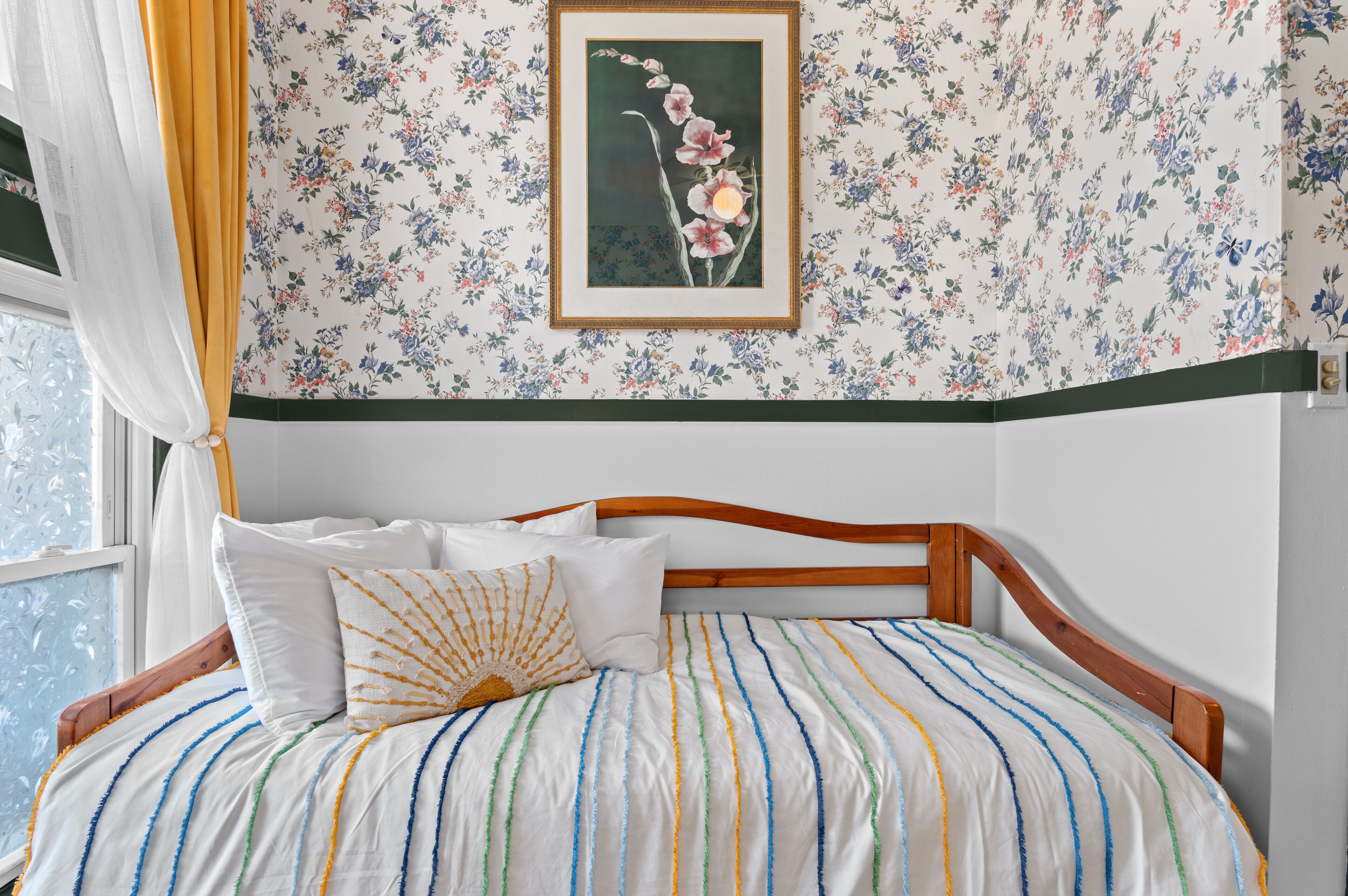 Quarto com cabeceira de madeira, cama com roupa de cama listrada, papel de parede floral e um quadro na parede.