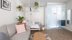 Uma sala de estar moderna com um sofá cinzento, uma mesa de centro de madeira e uma estante branca com plantas e livros.