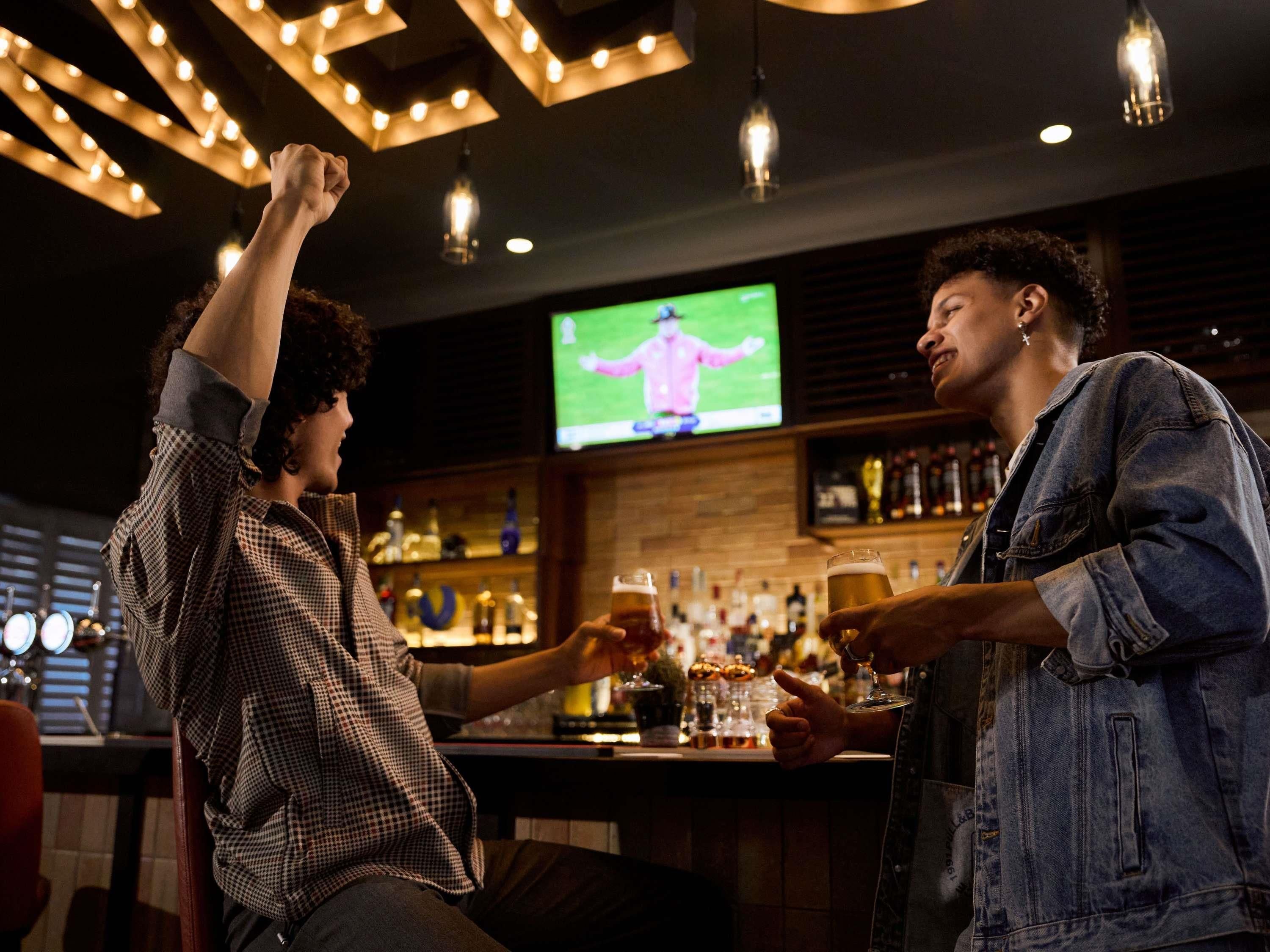 Um bar com uma televisão exibindo um jogo de esportes, balcão com copos e uma pessoa levantando a mão.