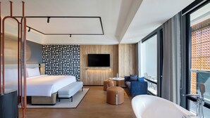 Quarto de hotel moderno com uma cama grande, área de estar, banheira e televisão na parede.