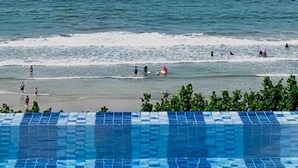 Vista de uma praia com pessoas nadando e surfando, e uma piscina em primeiro plano.