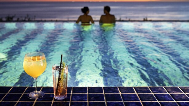 Piscina infinita com duas pessoas ao fundo, um copo de bebida com canudo e um raminho de alecrim na borda, e vista para o pôr do sol.