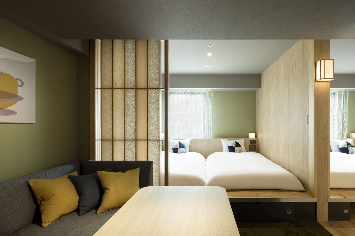 Quarto de hotel moderno com sofá, duas camas, divisória de madeira e vista pela janela.