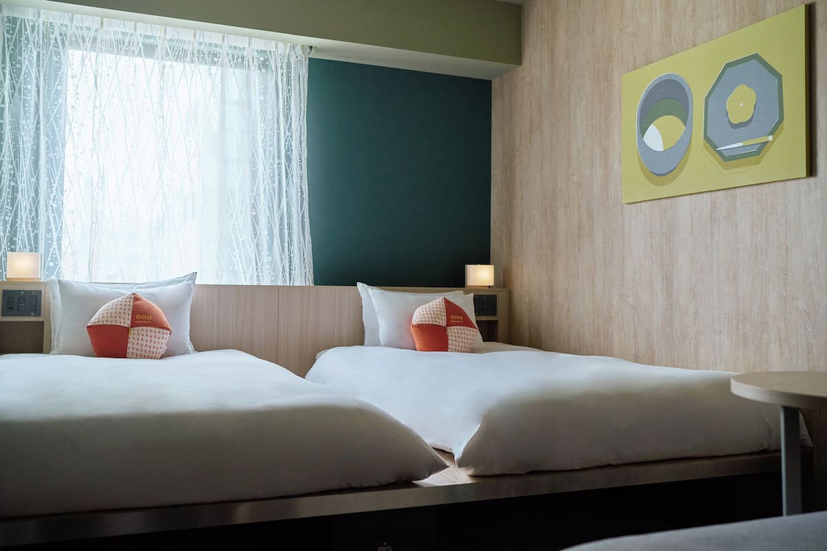 Quarto de hotel com duas camas, janela com cortinas translúcidas, uma obra de arte pendurada na parede e uma mesa de cabeceira com abajur.