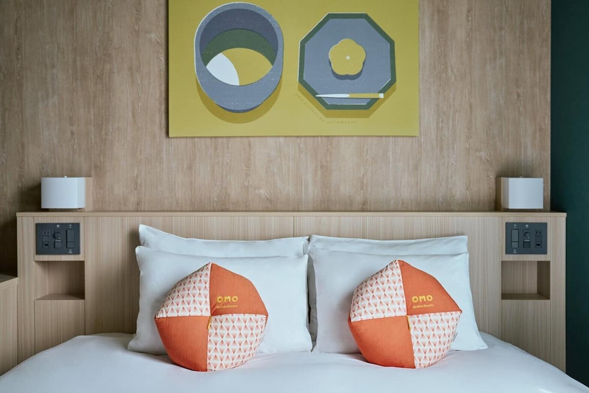 Quarto de hotel com cabeceira de madeira, duas almofadas com padrões geométricos, cama com roupa de cama branca e uma obra de arte pendurada na parede.