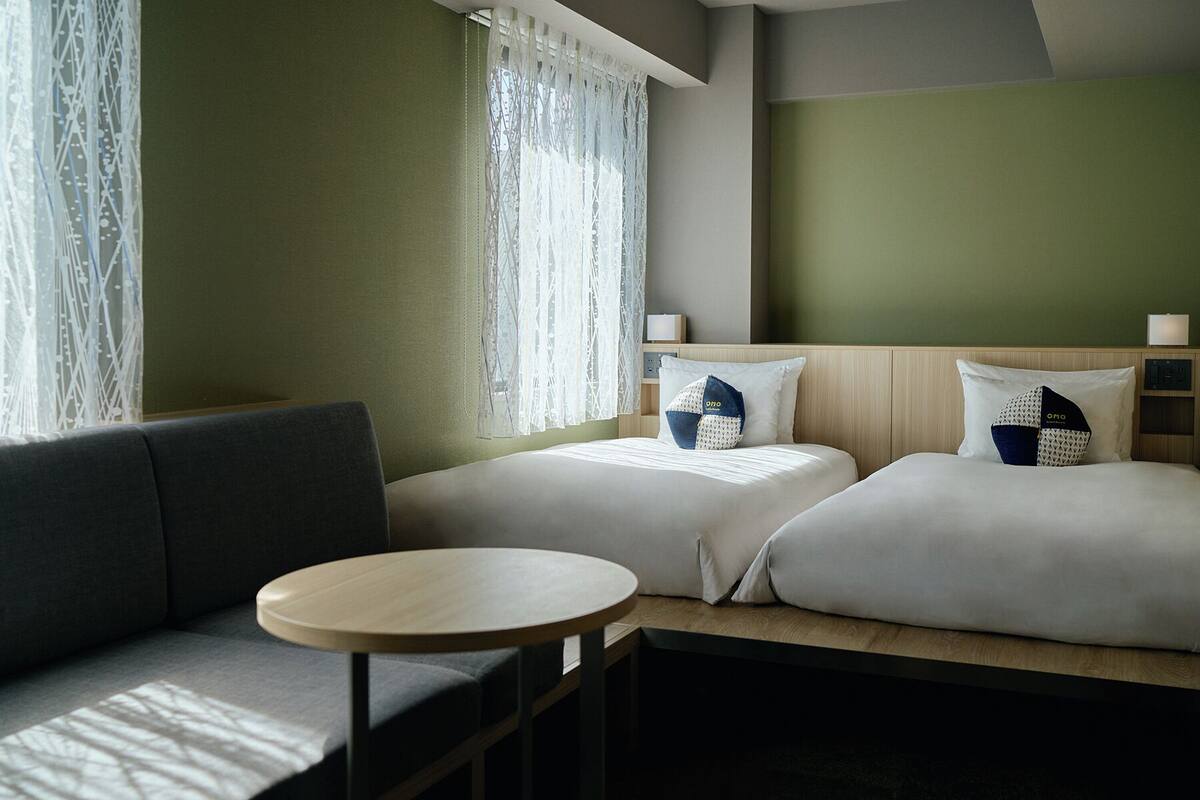 Quarto de hotel com duas camas, um sofá, uma mesa redonda pequena e uma janela com cortinas leves.