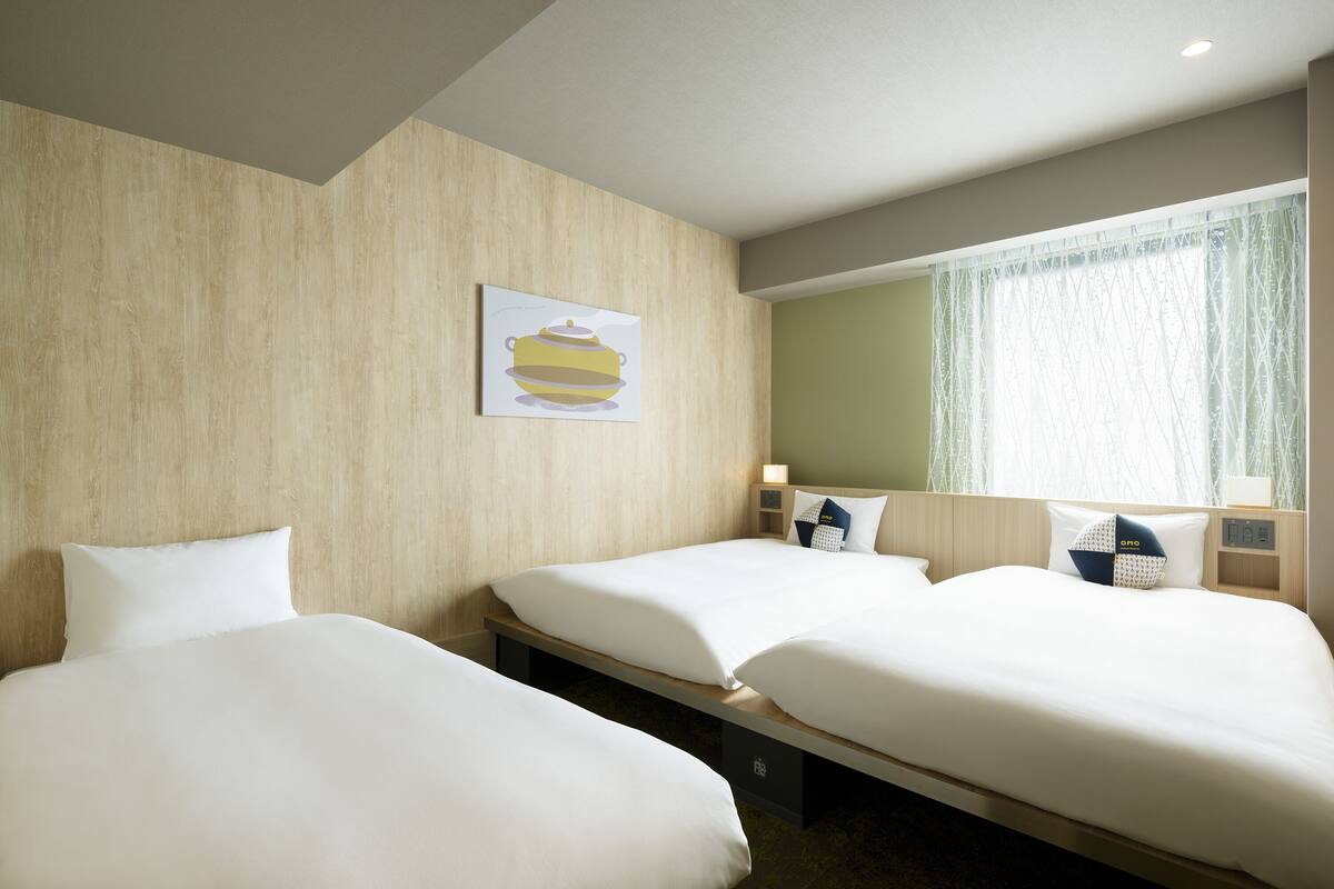 Quarto de hotel com duas camas, cabeceira de madeira, um quadro, mesa de cabeceira com abajur e janela com cortinas leves.