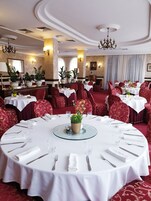 Uma sala de jantar formal com mesas redondas, cadeiras estofadas vermelhas e toalhas de mesa brancas.