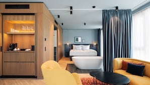 Quarto de hotel moderno com um guarda-roupa de madeira, uma cama grande, um sofá amarelo, uma mesa redonda e uma banheira.