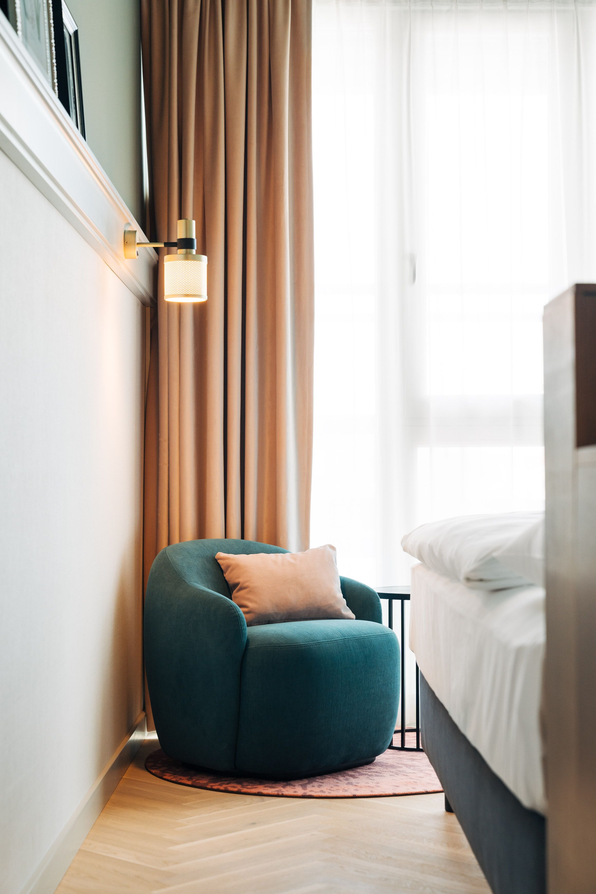 Quarto nos fundos com uma poltrona azul-esverdeada, uma cama, uma luminária na parede e uma janela com cortinas.