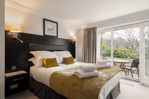 Quarto de hotel com uma cama grande, duas luminárias de cabeceira, um quadro na parede e uma varanda com mesa e cadeiras.