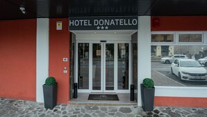 A entrada do Hotel Donatello, com fachada moderna, porta de vidro e área de estacionamento ao fundo.
