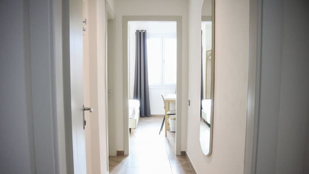 Corredor estreito que dá para um quarto com uma cama, uma cadeira e uma janela com cortinas.