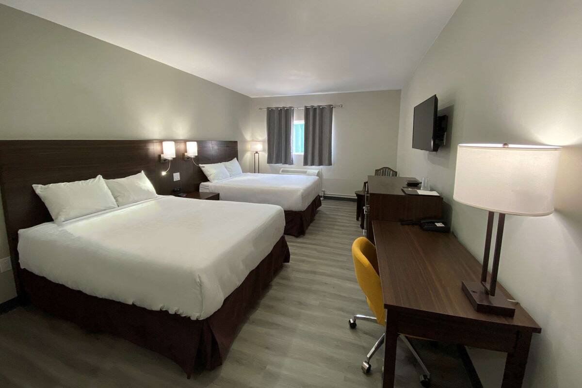 Quarto de hotel com duas camas, uma escrivaninha, uma cadeira, televisão e janela com cortinas.