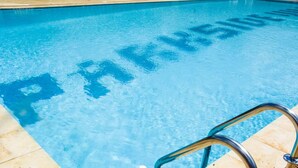 Uma piscina azul e límpida com uma escada de metal que dá acesso à água, rodeada por um deck revestido com azulejos.