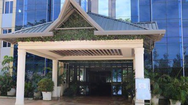 Uma entrada coberta com telhado triangular, plantas em vasos e um edifício moderno com janelas de vidro refletivas ao fundo.