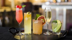 Uma seleção de cocktails numa bandeja, incluindo uma bebida rosa com morangos, uma bebida amarela com hortelã, uma bebida verde com pepino e uma bebida transparente com uma fatia de limão.