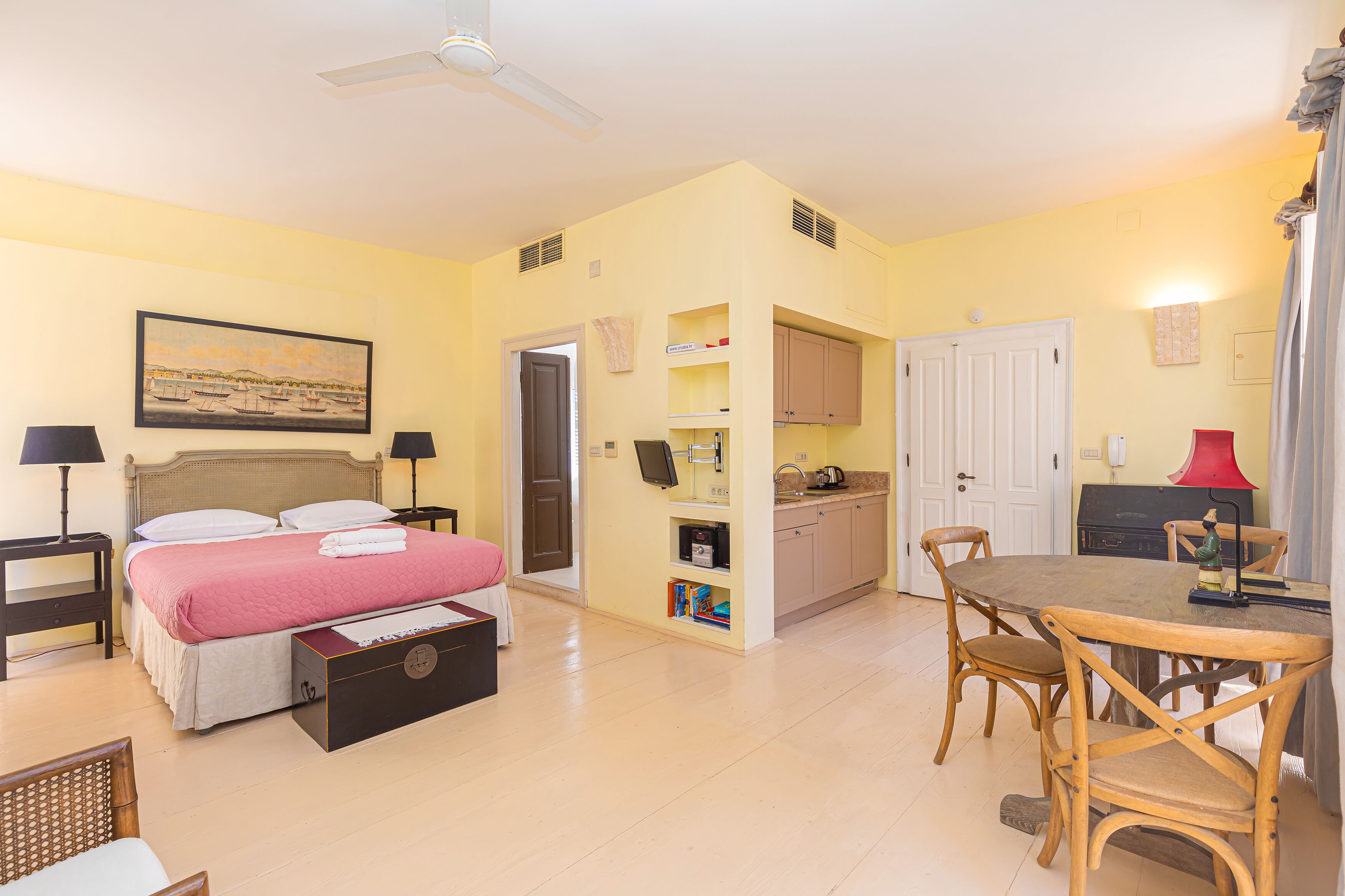 Quarto com cama, mesas de cabeceira, escrivaninha e uma cadeira. Há uma porta que dá para o banheiro e uma área de cozinha.